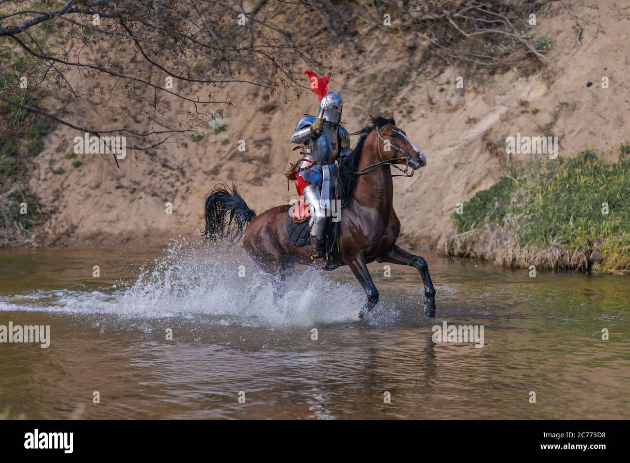 A young adult man in knightly armor rides a horse on a river along a ...
