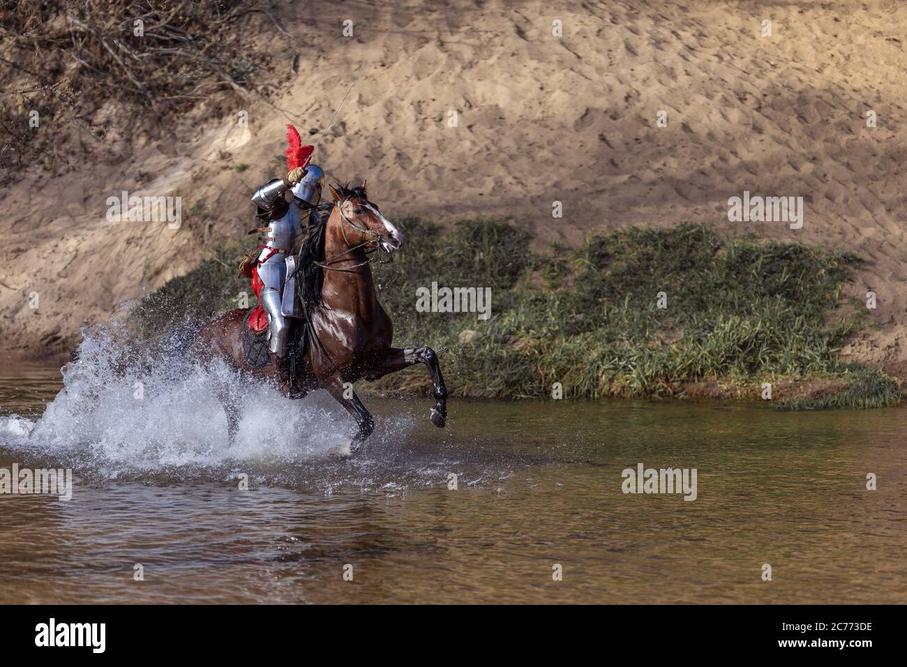 A young adult man in knightly armor rides a horse on a river along a ...