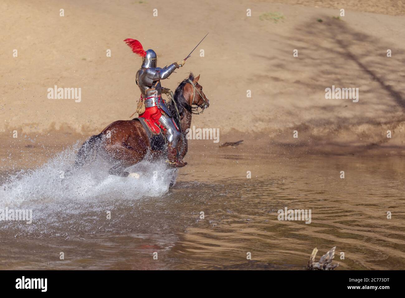 A young adult man in knightly armor rides a horse on a river along a ...