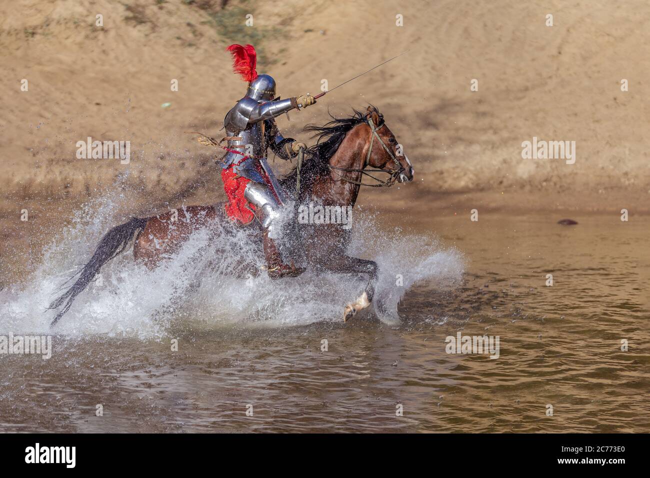A young adult man in knightly armor rides a horse on a river along a ...