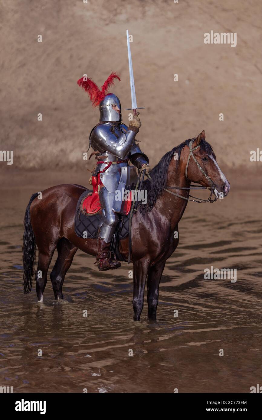 A young adult man in knightly armor rides a horse on a river along a ...