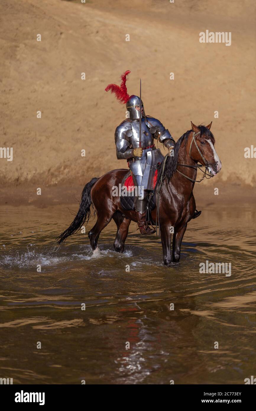 A young adult man in knightly armor rides a horse on a river along a ...