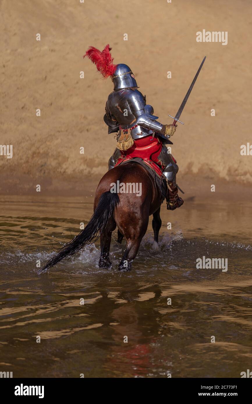 A young adult man in knightly armor rides a horse on a river along a ...