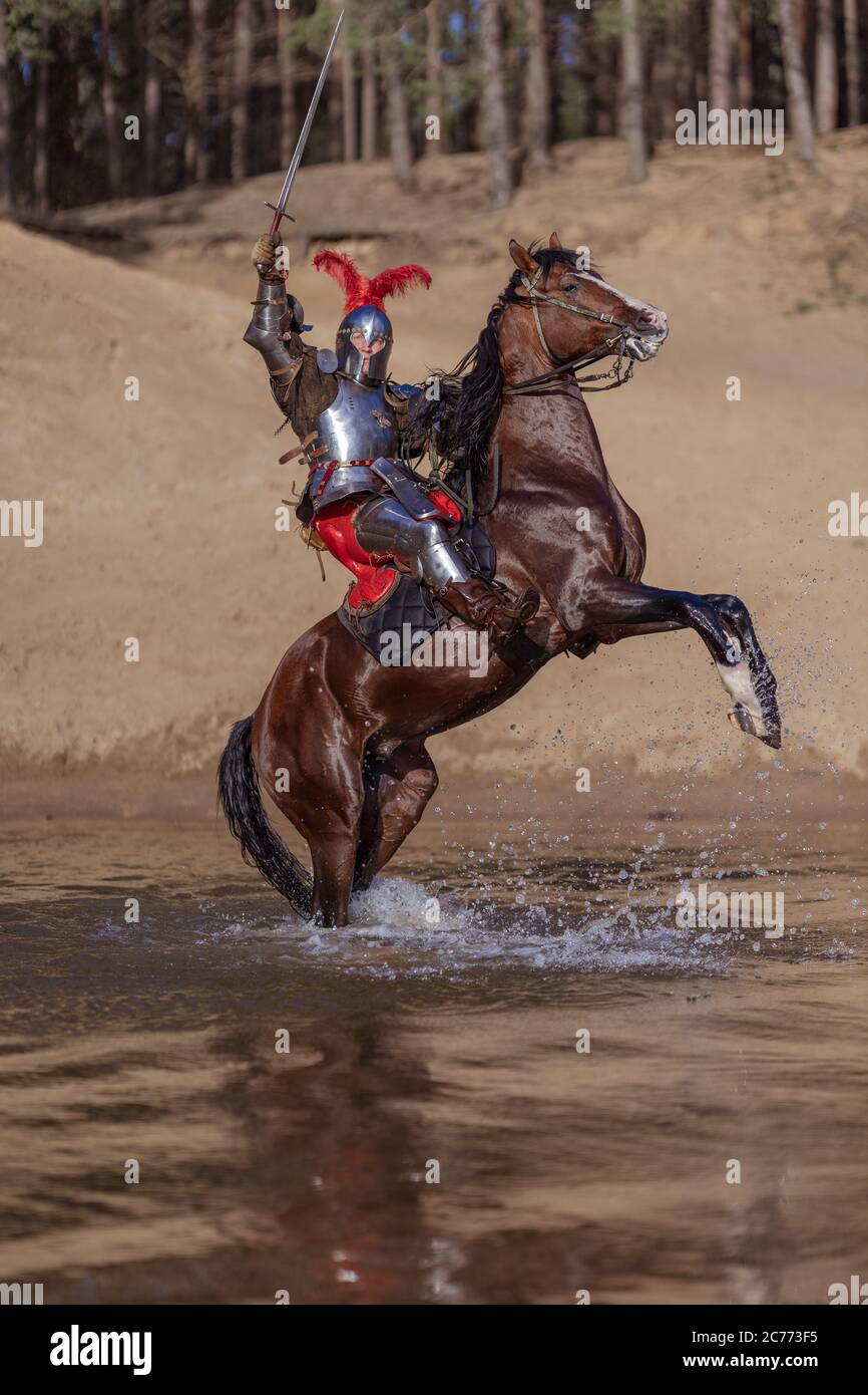 A young adult man in knightly armor rides a horse on a river along a ...