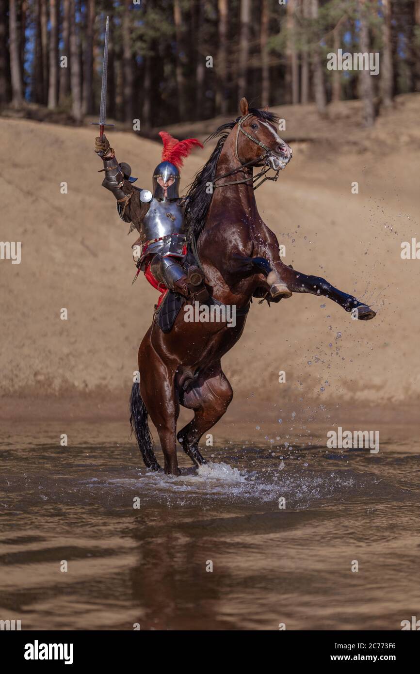 A young adult man in knightly armor rides a horse on a river along a ...