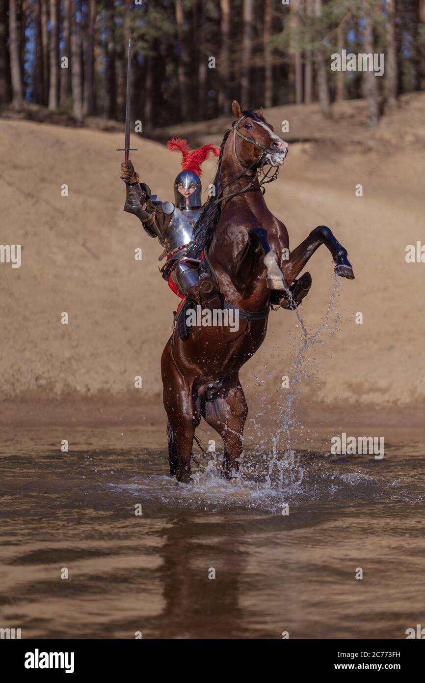 A young adult man in knightly armor rides a horse on a river along a ...