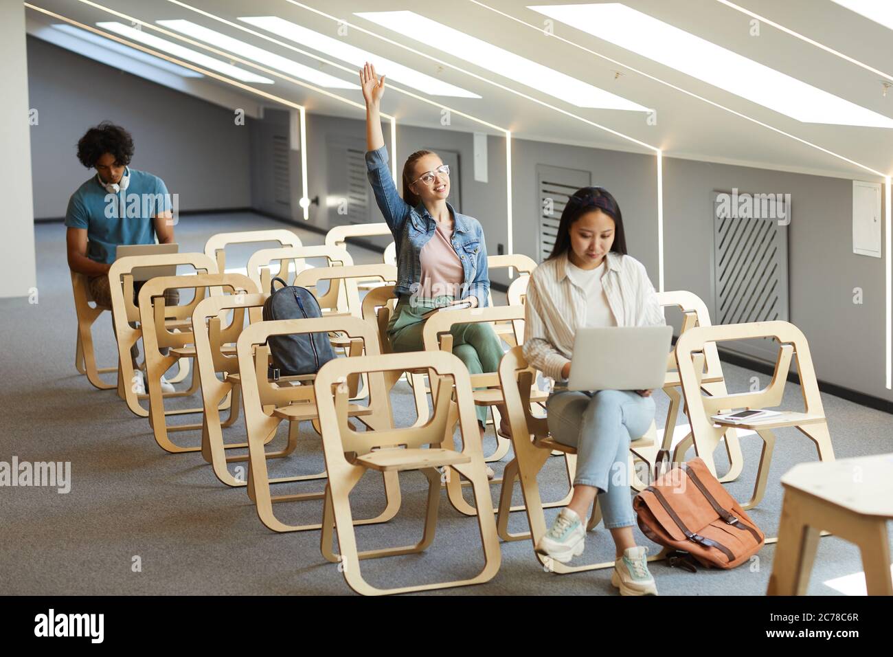 Curious student girl sitting on wooden chair and raising arm while ...