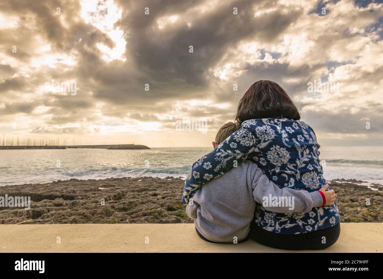 Mom with son sitting and hugging each other on the wall facing the sea ...