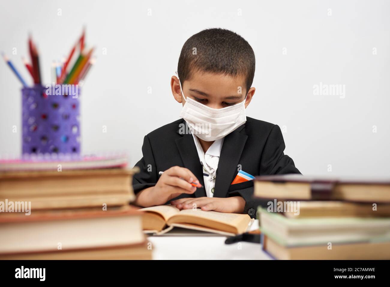 Young schoolboy in face mask doing homework between pile of books ...