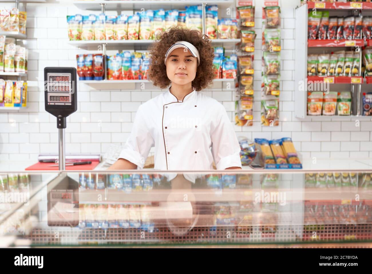 Front view of cheerful girl posing behind glass store counter, shelfs ...