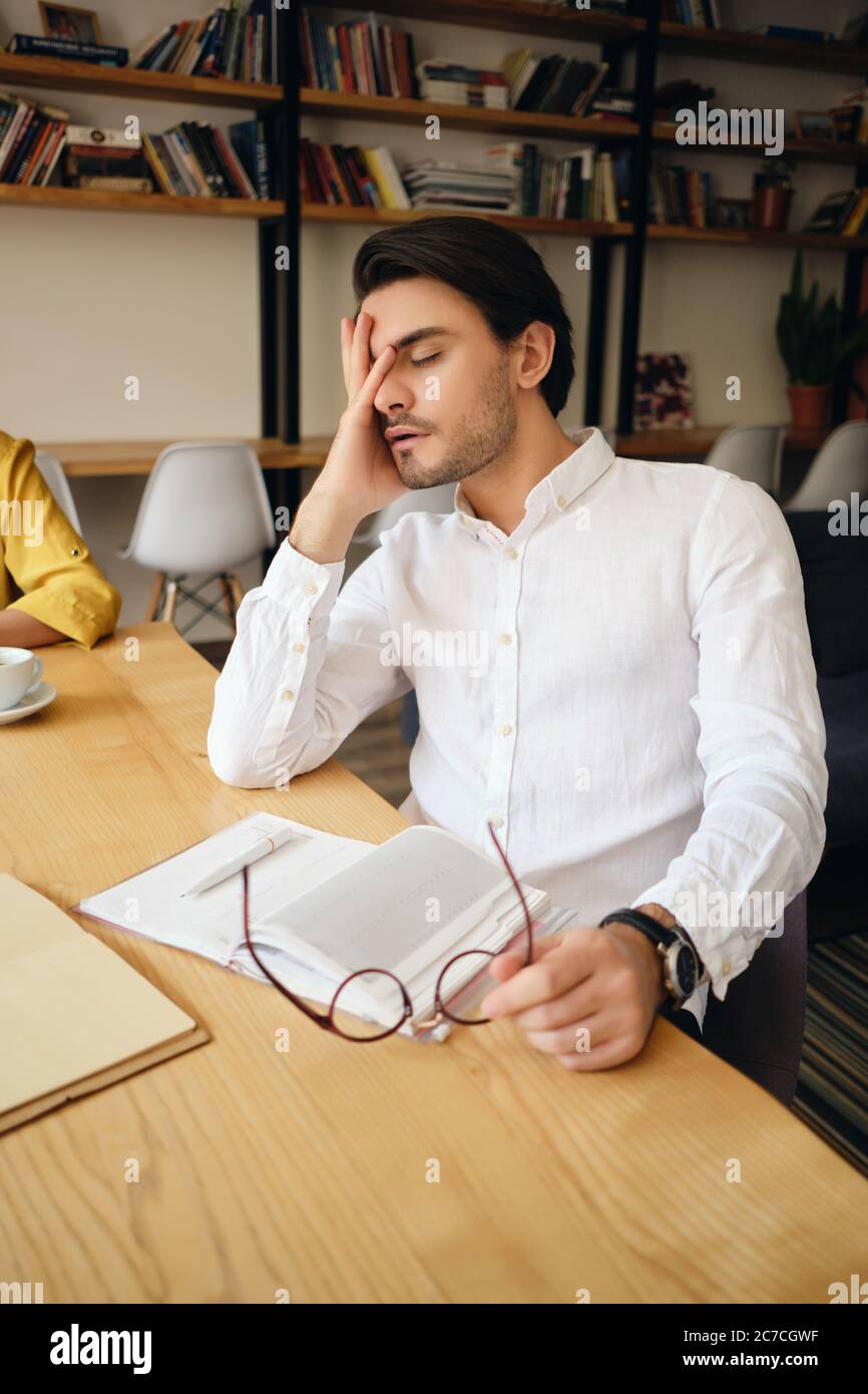 Young tired man sitting at the table with notepad leaning head on hand ...
