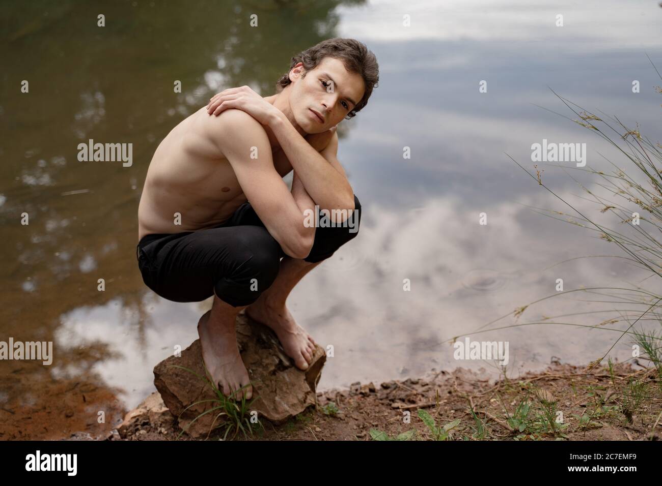 Thoughtful Shirtless Man Crouching On Rock At Lakeshore In Forest Stock