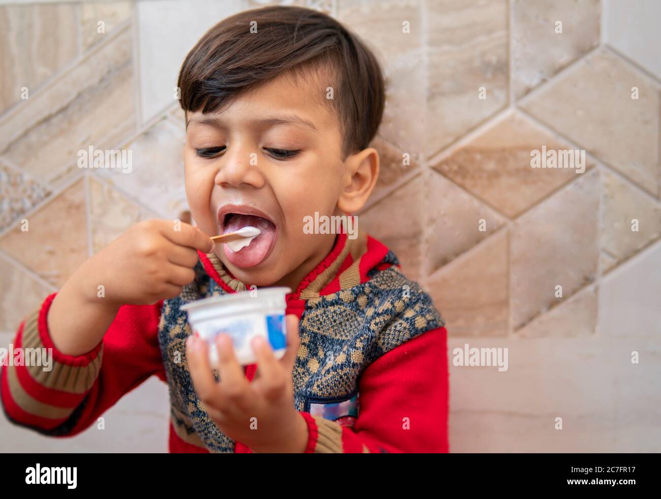 Cute little boy in red dress, eating cup ice cream with spoon at home ...