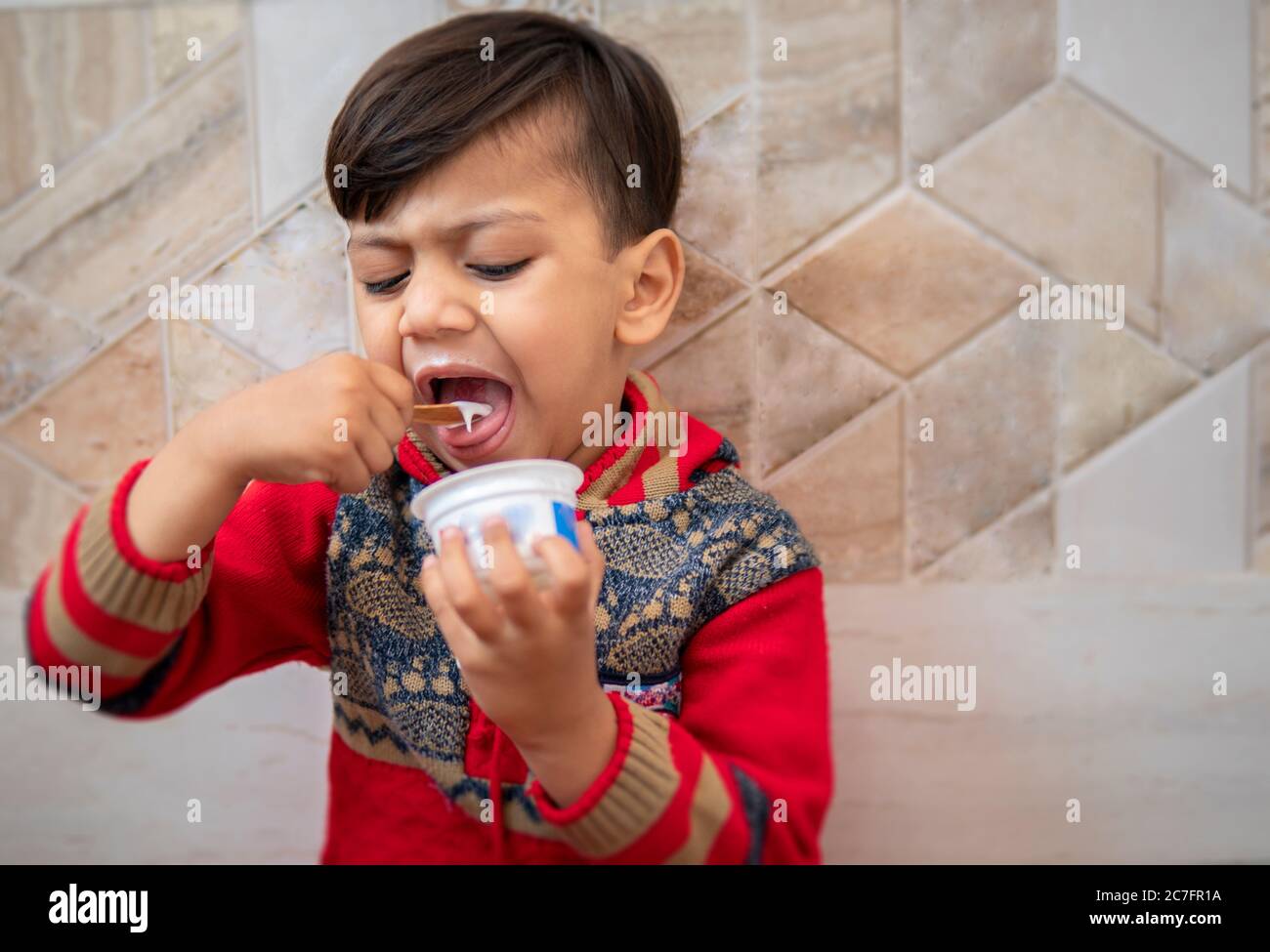 Cute little boy in red dress, eating cup ice cream with spoon at home ...