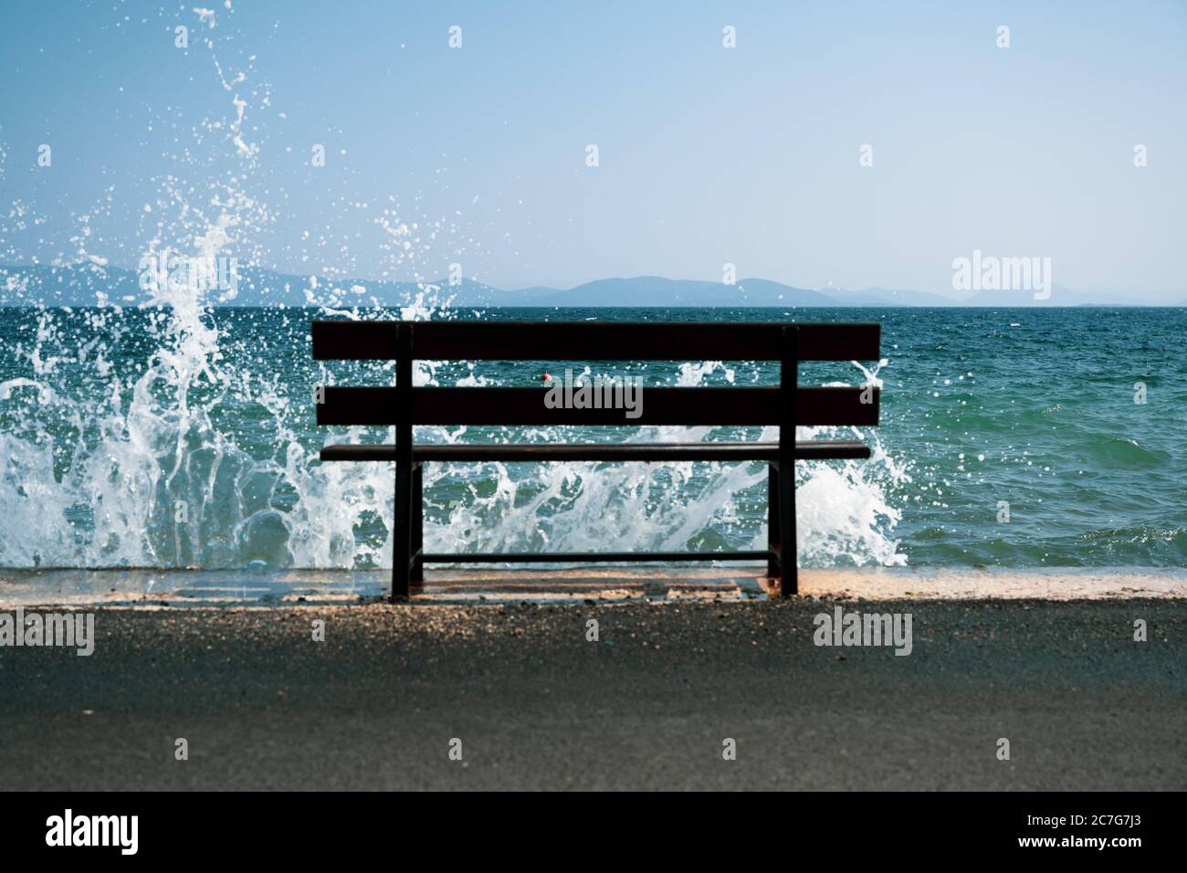 A dark park bench stands at a pier where waves break and spray jumps up ...