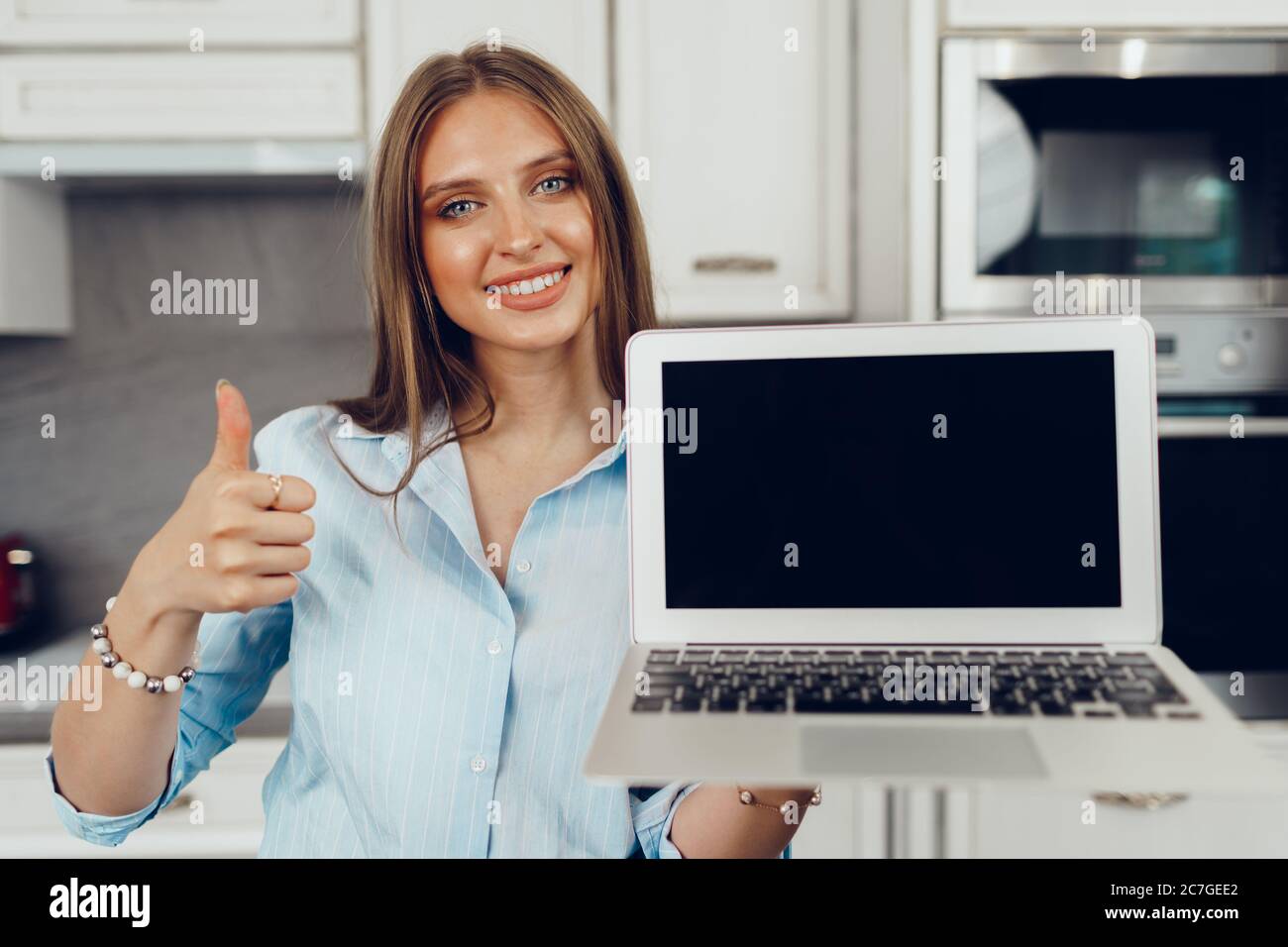 Young blonde woman standing in kithen and showing blank screen laptop ...