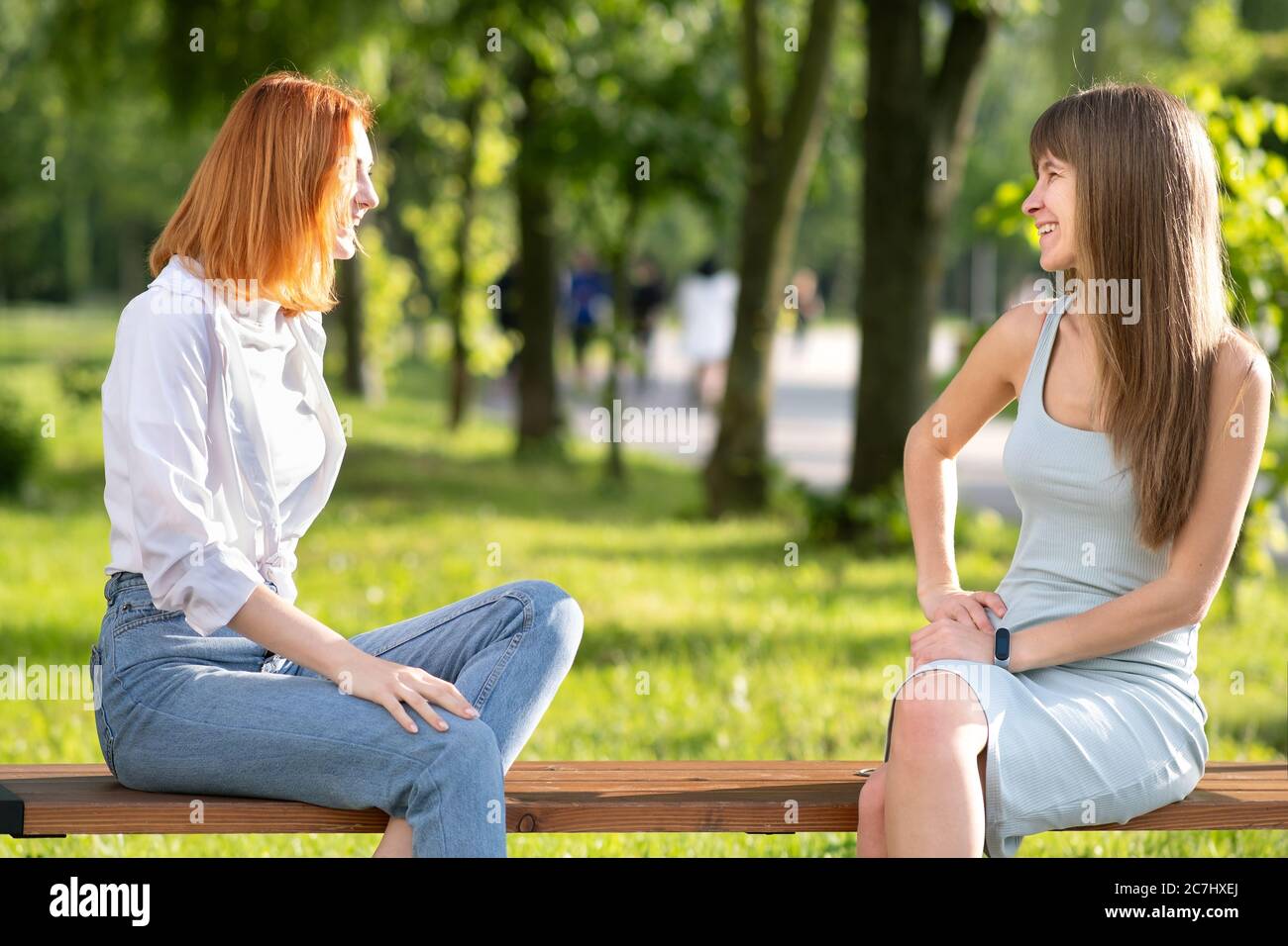 Two Young Girls Friends Sitting On A Bench In Summer Park Chatting Happily Having Fun Stock