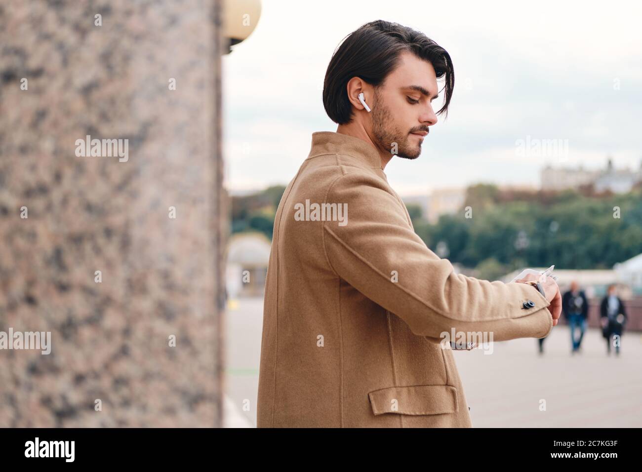 Side view of young serious man in beige coat watching time on ...