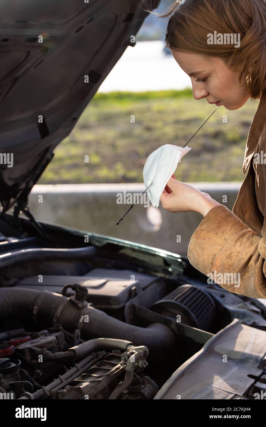 Confident caucasian woman driver checking oil level in car engine dipstick, cityscape on ...
