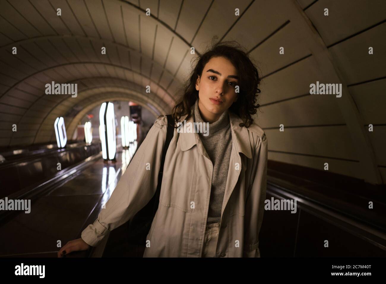 Beautiful girl in trench coat dreamily looking in camera on escalator ...