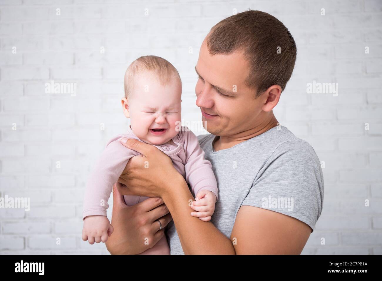 portrait of young father holding crying baby girl over white brick wall ...