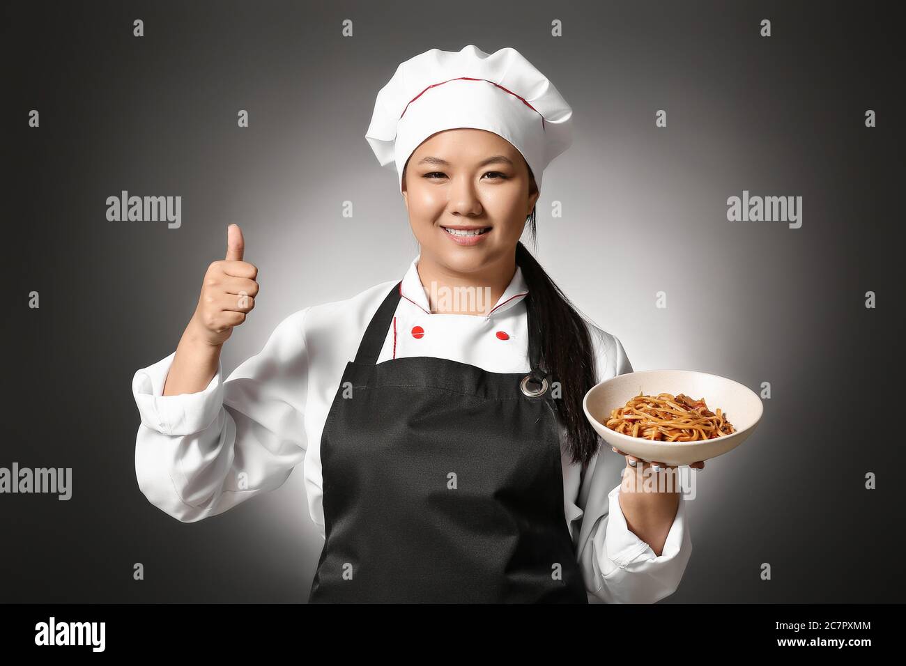 Beautiful Asian chef with dish showing thumb-up gesture on dark ...