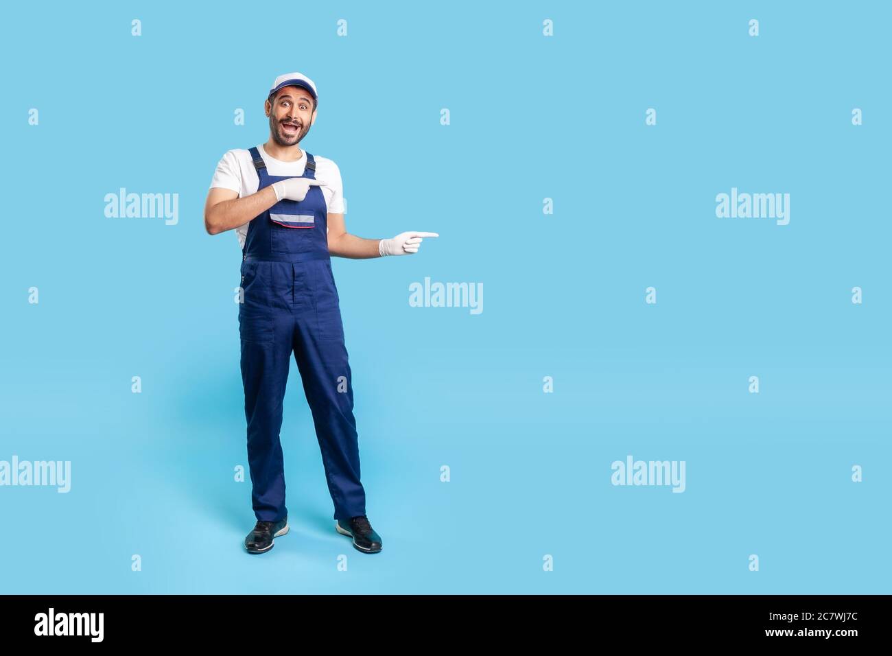 Full length surprised handyman in uniform pointing at copy space ...