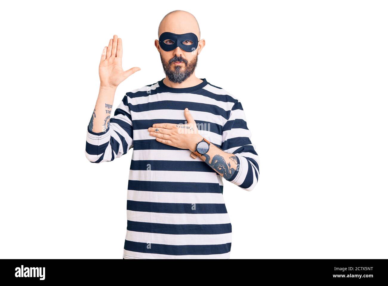 Young handsome man wearing burglar mask swearing with hand on chest and ...
