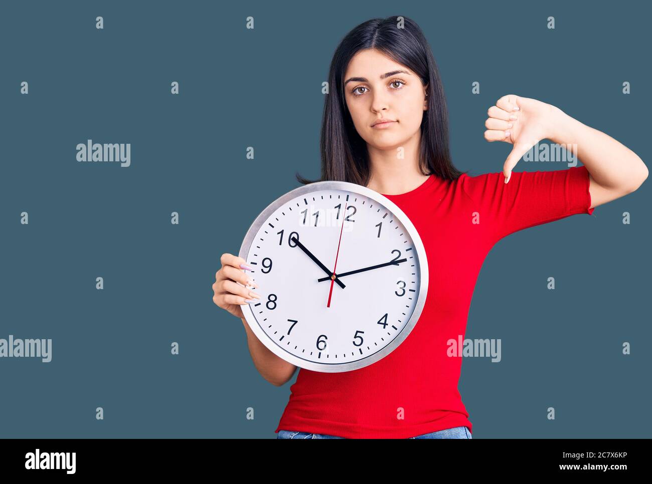 Young beautiful girl holding clock with angry face, negative sign ...