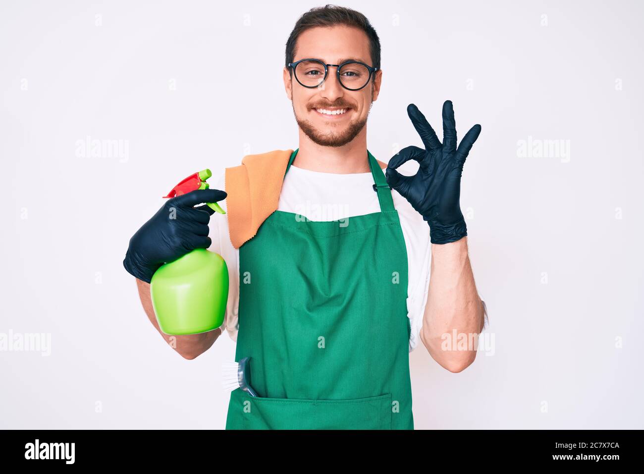 Young handsome man wearing apron holding sprayer doing ok sign with ...