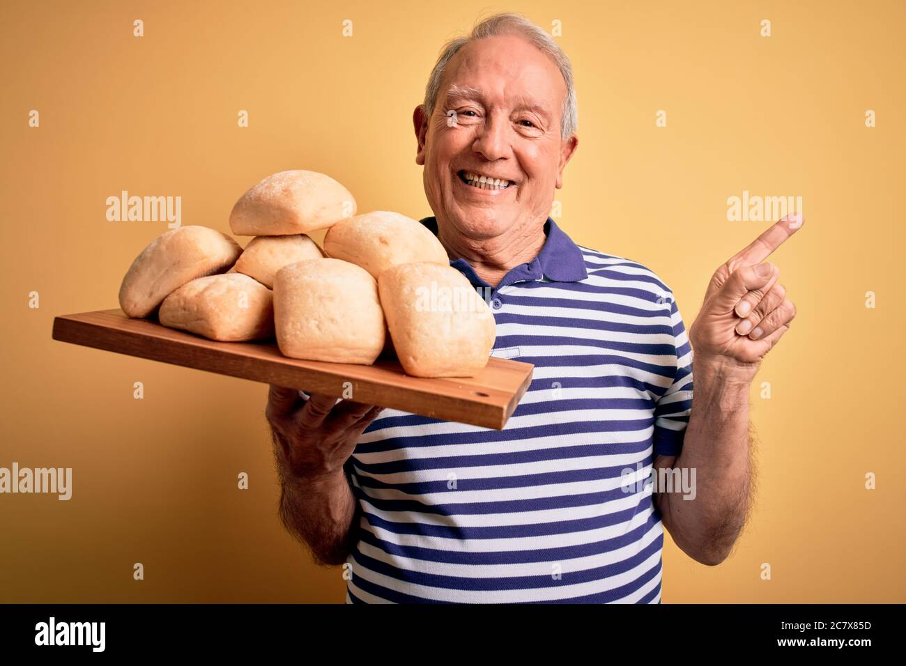 Senior grey haired baker man holding fresh homemade bread over yellow ...