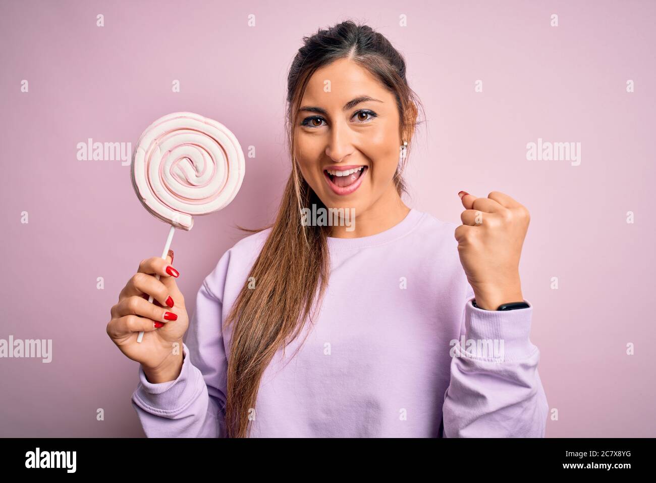 Young beautiful brunette woman eating sweet candy over isolated pink ...