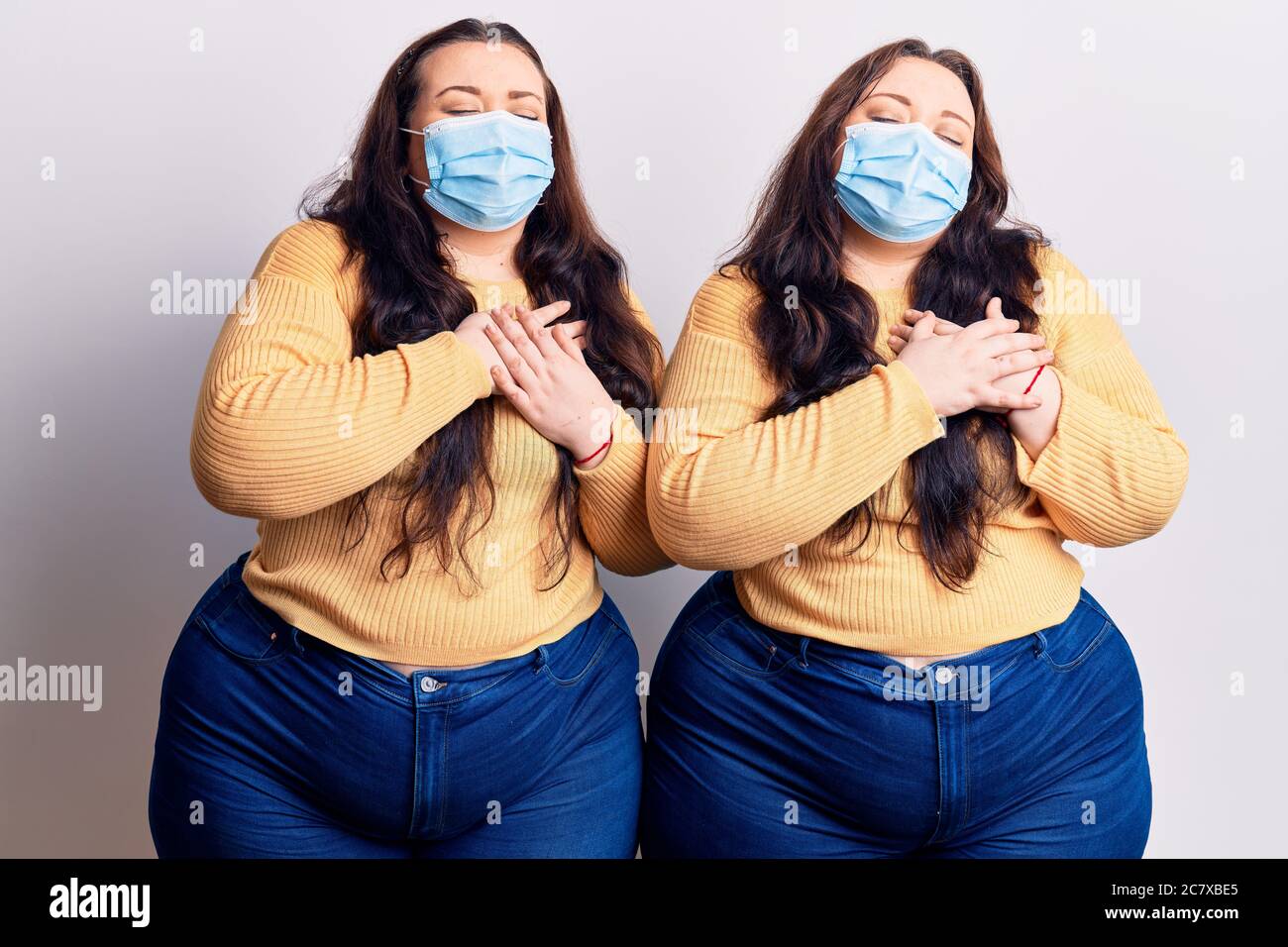 Young plus size twins wearing medical mask smiling with hands on chest ...