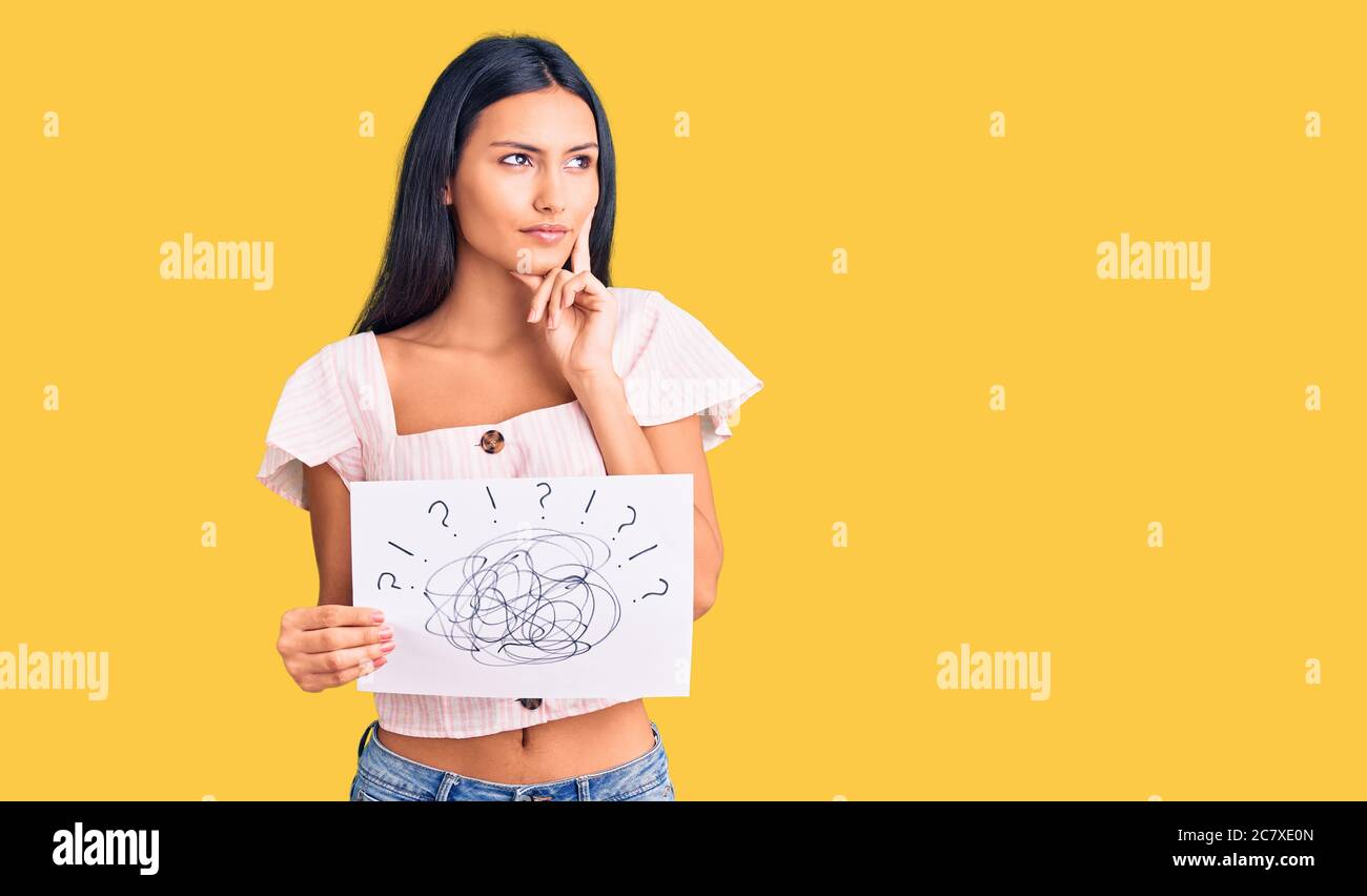 Young beautiful latin girl holding scribble draw serious face thinking about question with hand ...