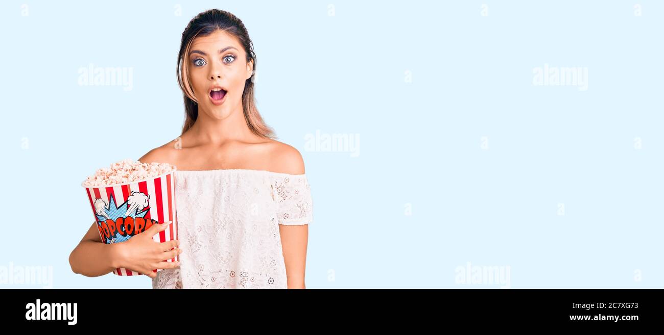 Young beautiful woman holding popcorn scared and amazed with open mouth ...