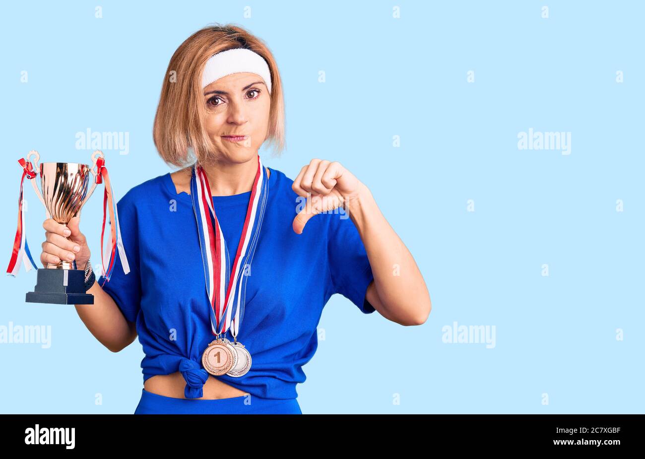 Young blonde woman holding champion trophy wearing medals with angry ...