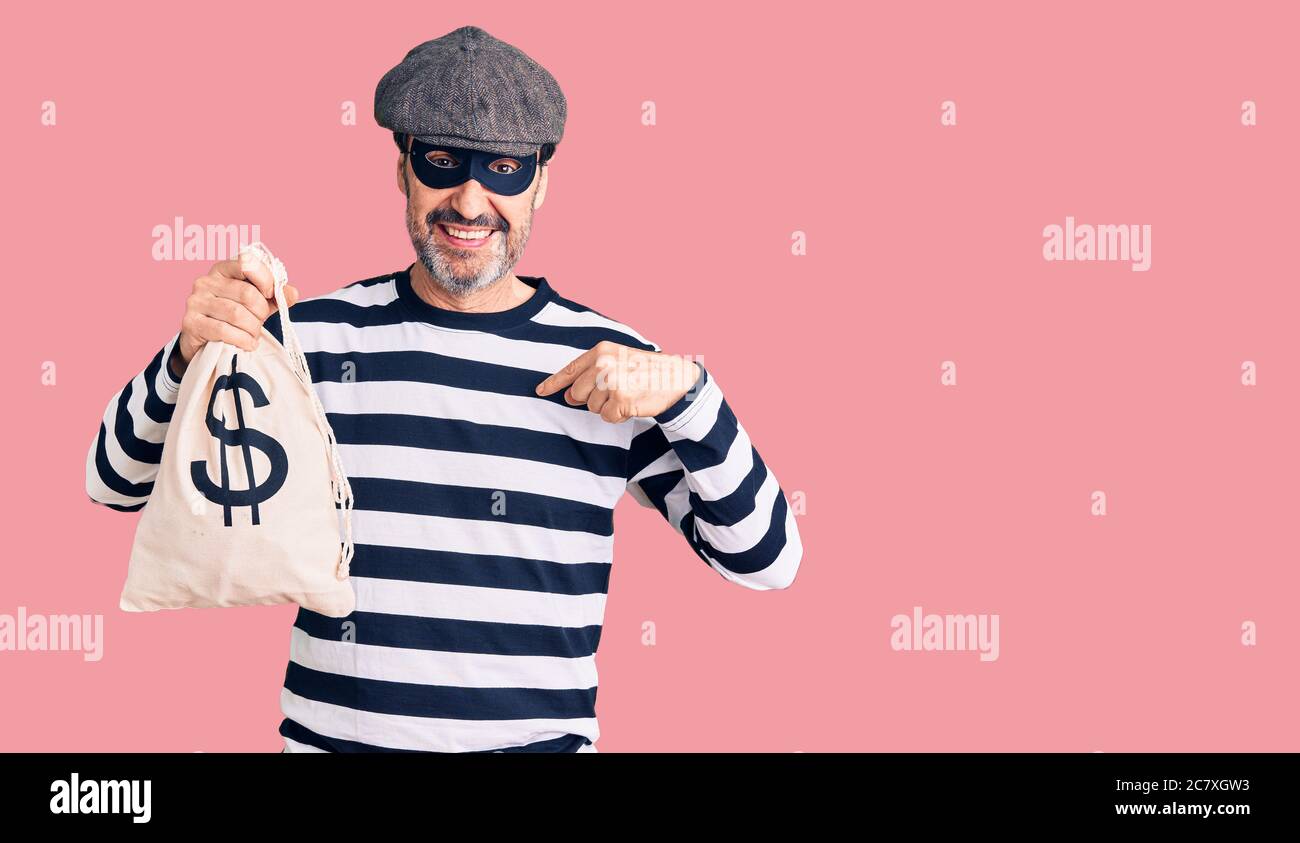 Middle age handsome man wearing burglar mask holding money bag pointing ...