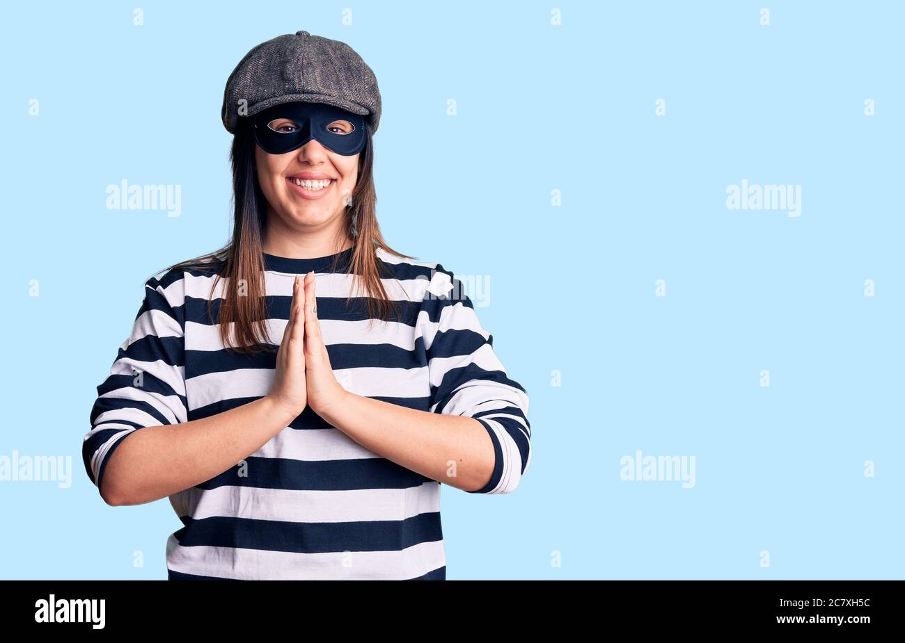 Young Beautiful Brunette Woman Wearing Burglar Mask Praying With Hands