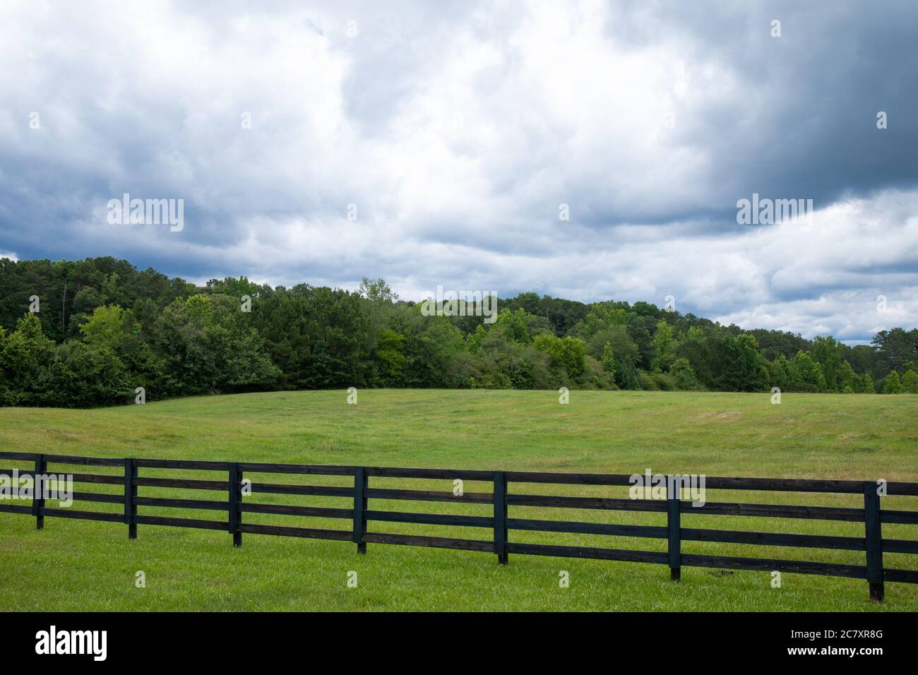 Storm clouds over a brilliant green field, black rail fence in front ...