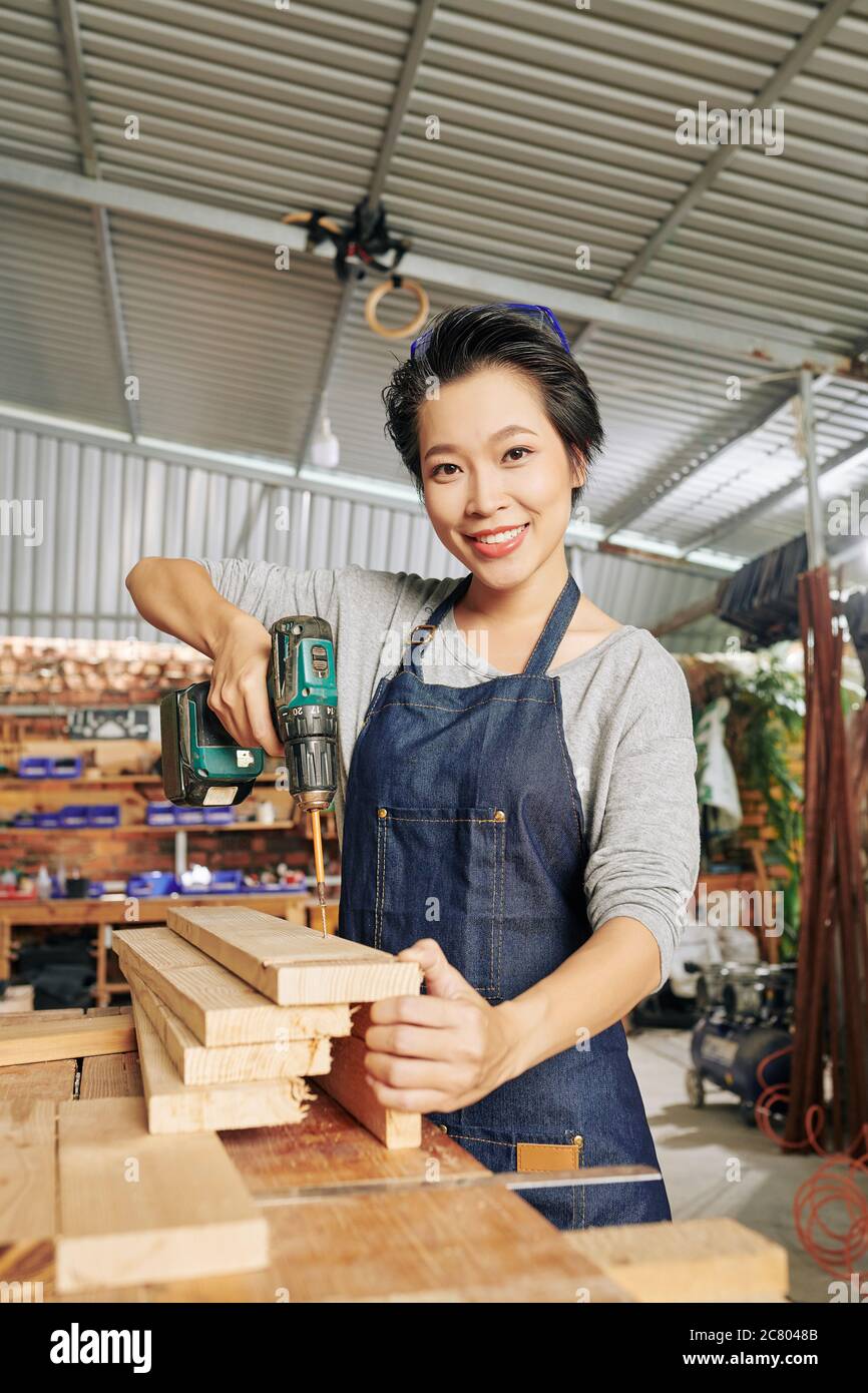 Portrait of smiling female Asian carpenter in denim apron drilling ...