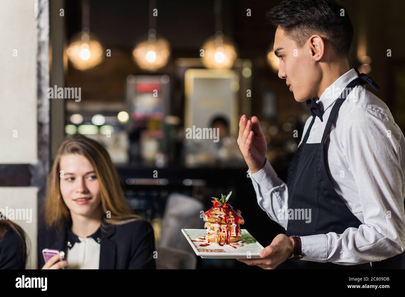 Positive young waiter serving terrace restaurant guests at table.Focus ...