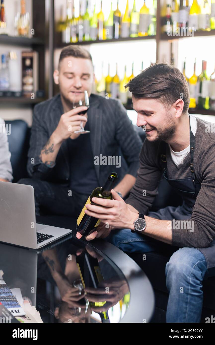Focused young male sommelier in suite looking at red wine in bottle ...