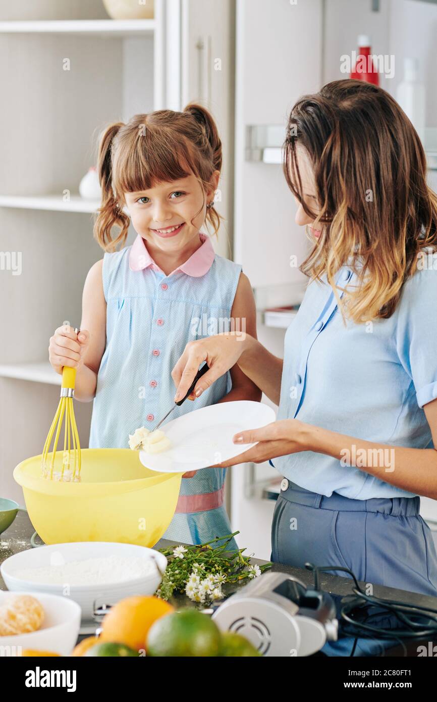 Young woman adding soft butter in big plastic bowl when her daughter ...