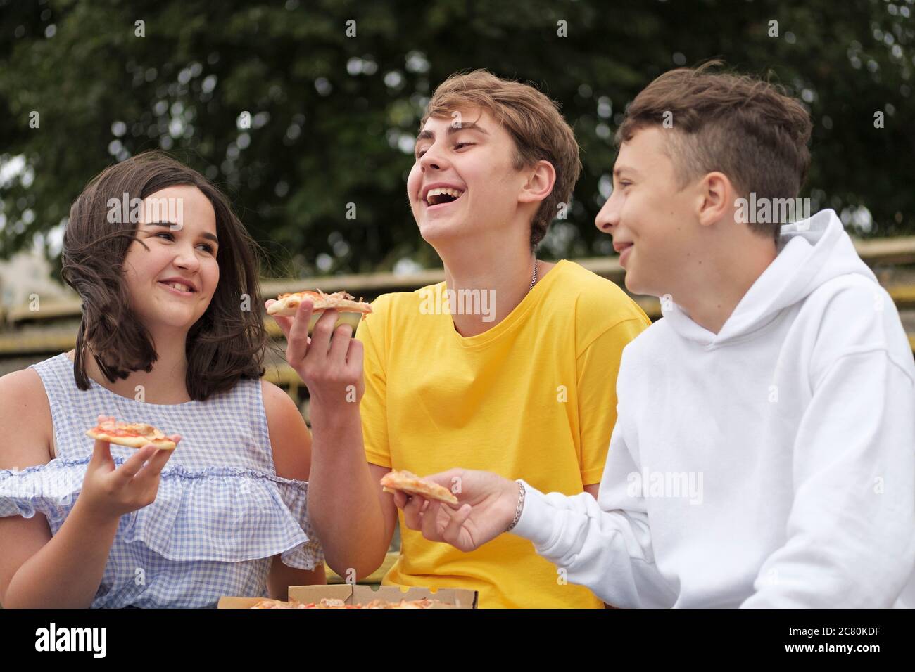 Two teenage boys and a girl sharing snacks outdoors laughing and joking ...
