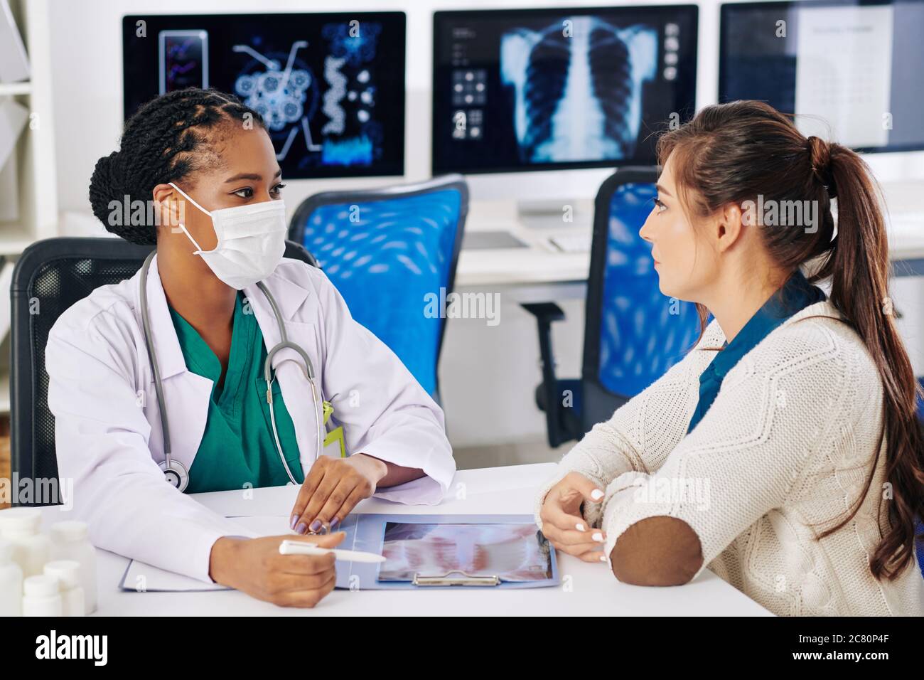 Black female general practitioner in medical mask discussing x-ray with ...