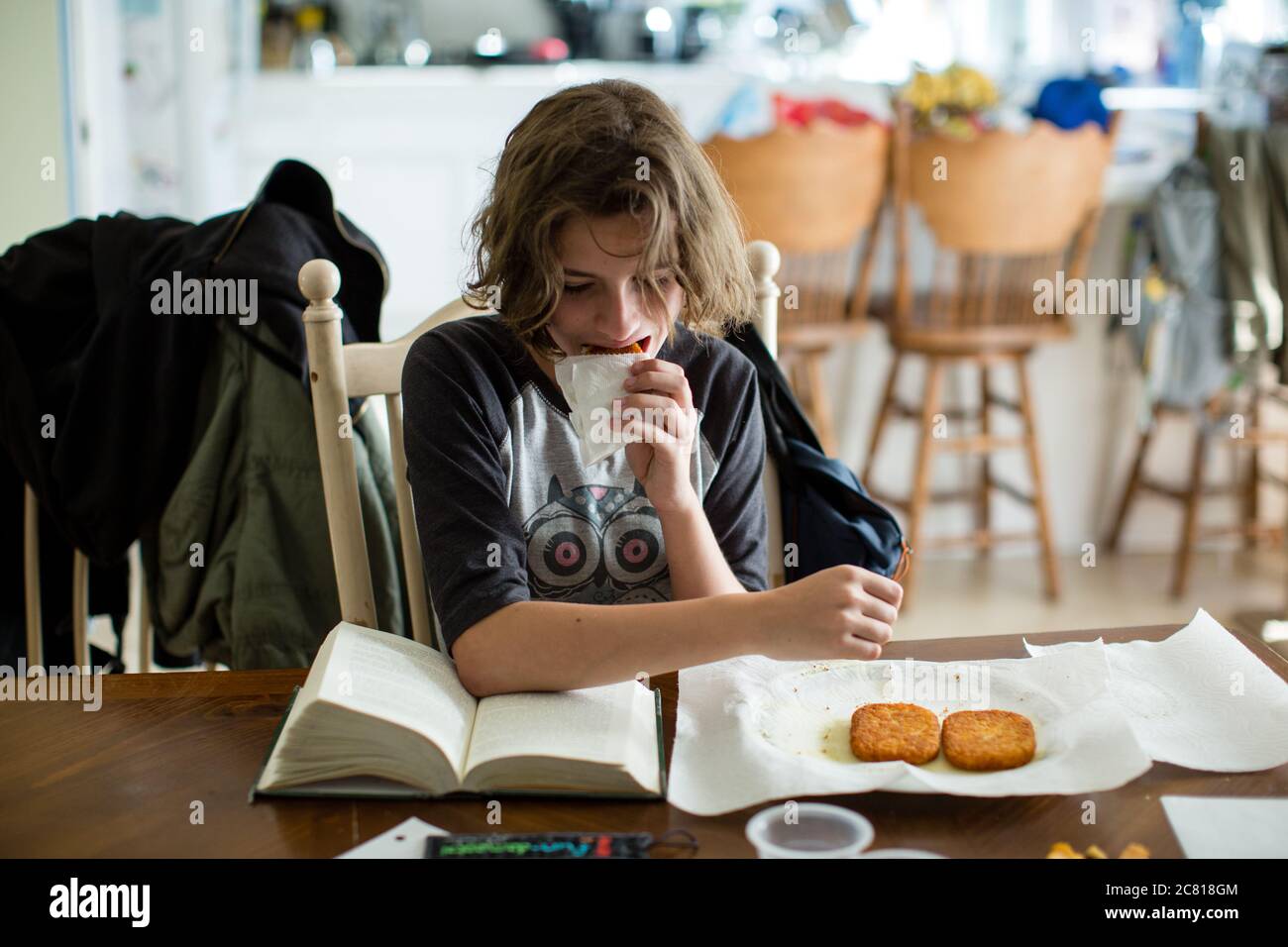 Teen girl takes a bite of a hash brown while she reads her book Stock ...