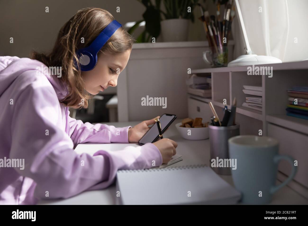 Focused teenage girl wearing headphones holding smartphone, writing ...