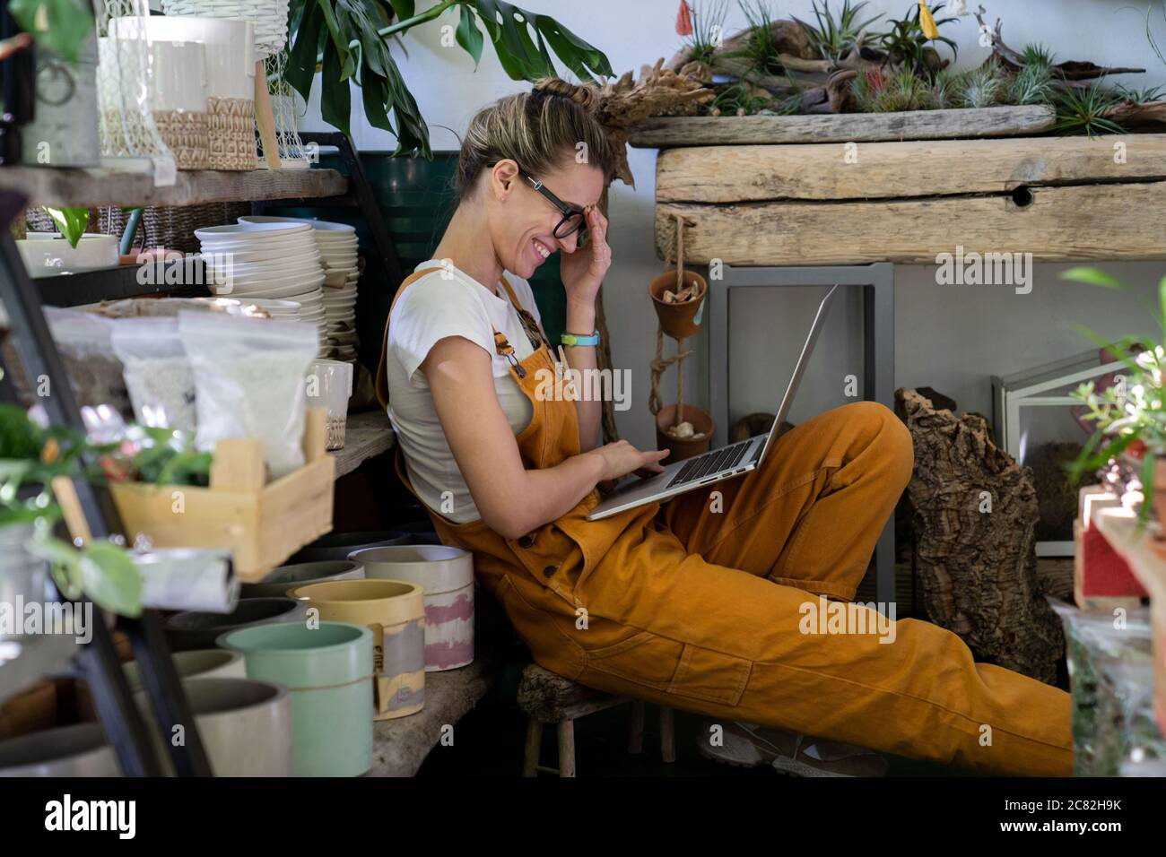 Cute woman gardener in glasses wear orange overalls, sitting on the floor in flower shop using ...