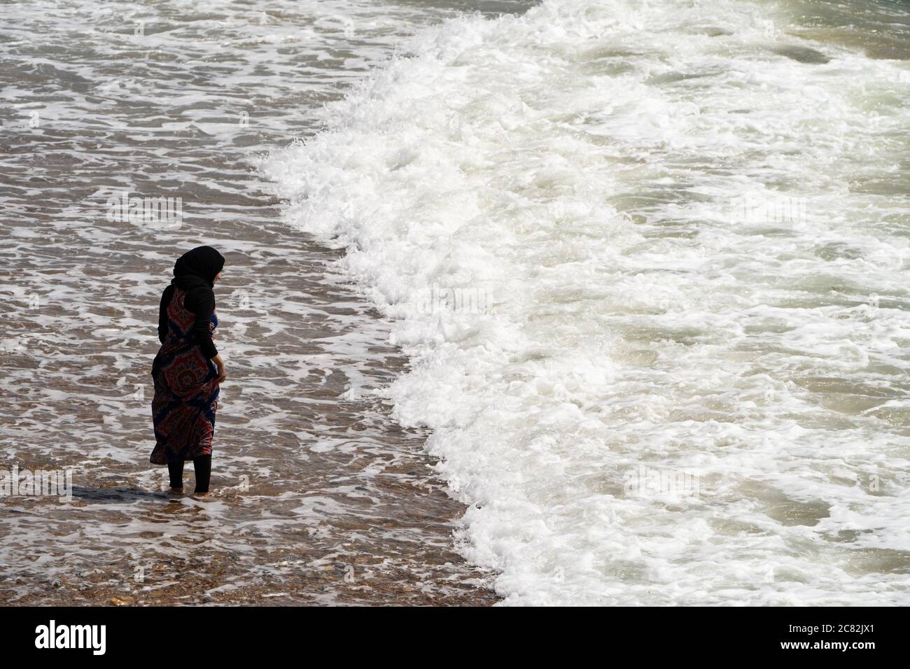 A young asian girl, enjoying paddling in the sea on a summer day at the seaside, fully clothed ...