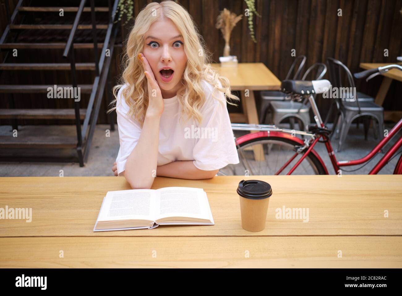 Attractive shocked blond girl amazedly looking in camera with book and ...